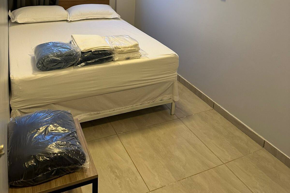 Cama bem arrumada com travesseiros e um cobertor dobrado, cabeceira de madeira, uma pequena mesa com um saco plástico e uma janela com cortinas.