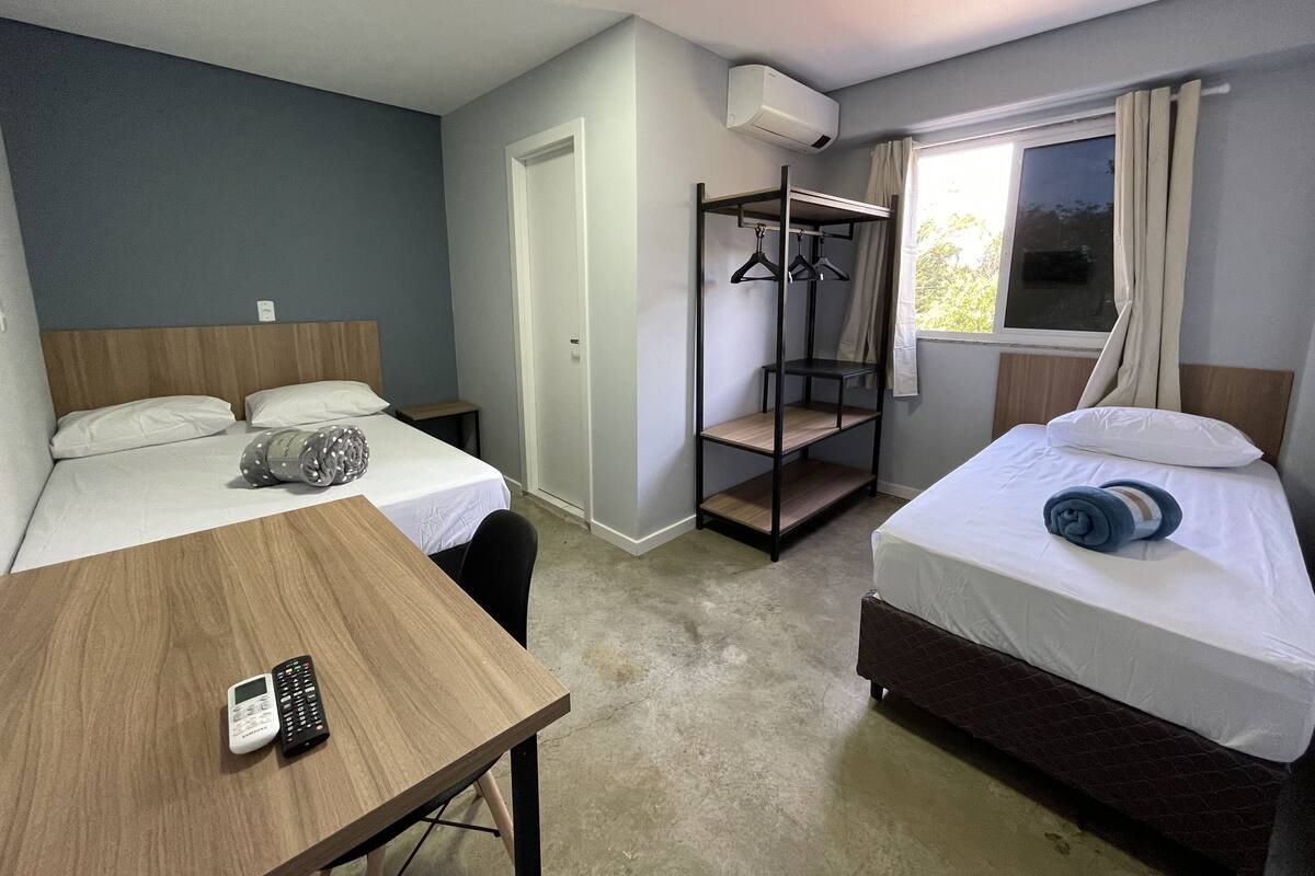 Quarto de hotel pequeno com duas camas, uma mesa com um controle remoto, uma cama beliche com um porta-roupas e uma janela com cortinas.