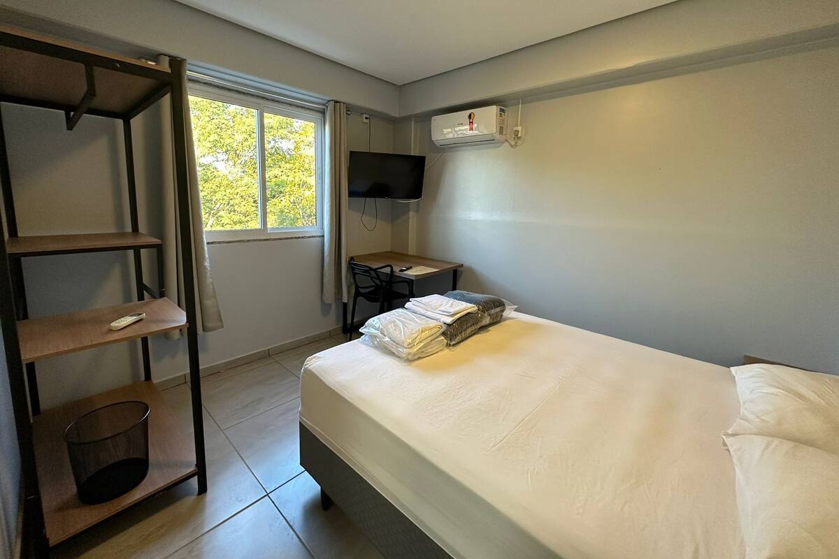 Quarto de hotel compacto com cama, escrivaninha, televisão e janela com vista para árvores.