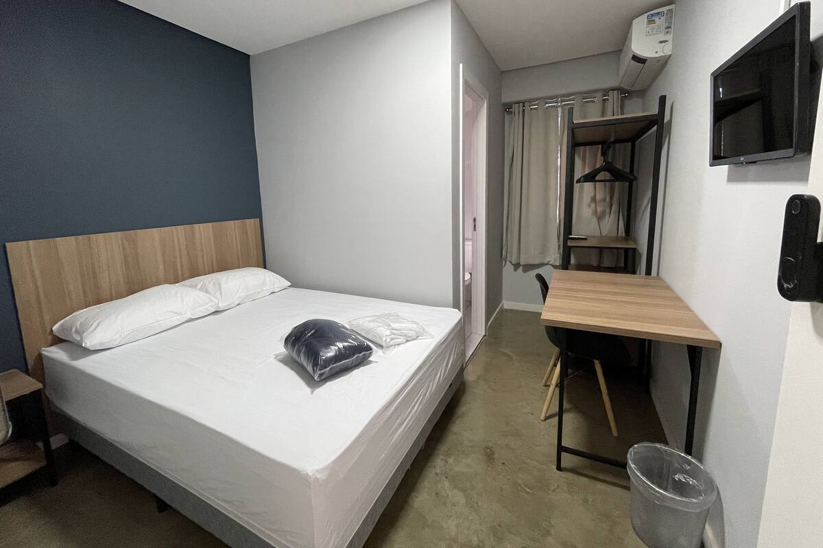 Quarto de hotel compacto com uma cama de solteiro, uma pequena escrivaninha, uma televisão e um guarda-roupa.