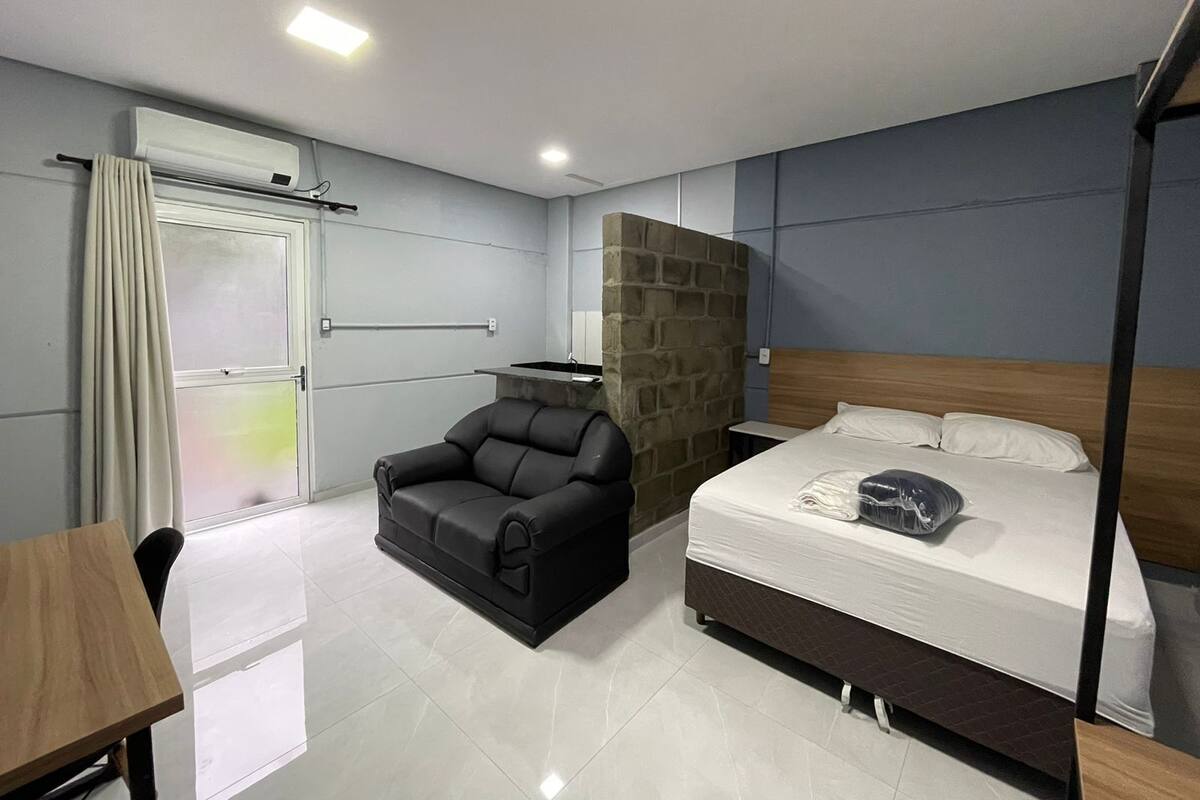 Quarto de hotel moderno com uma cama, uma poltrona de couro preto, uma mesinha e uma parede com detalhes em pedra.