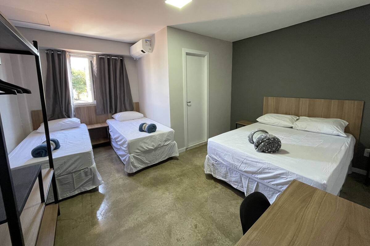 Quarto de hotel com duas camas de solteiro, uma escrivaninha de madeira, uma cadeira e um guarda-roupa.