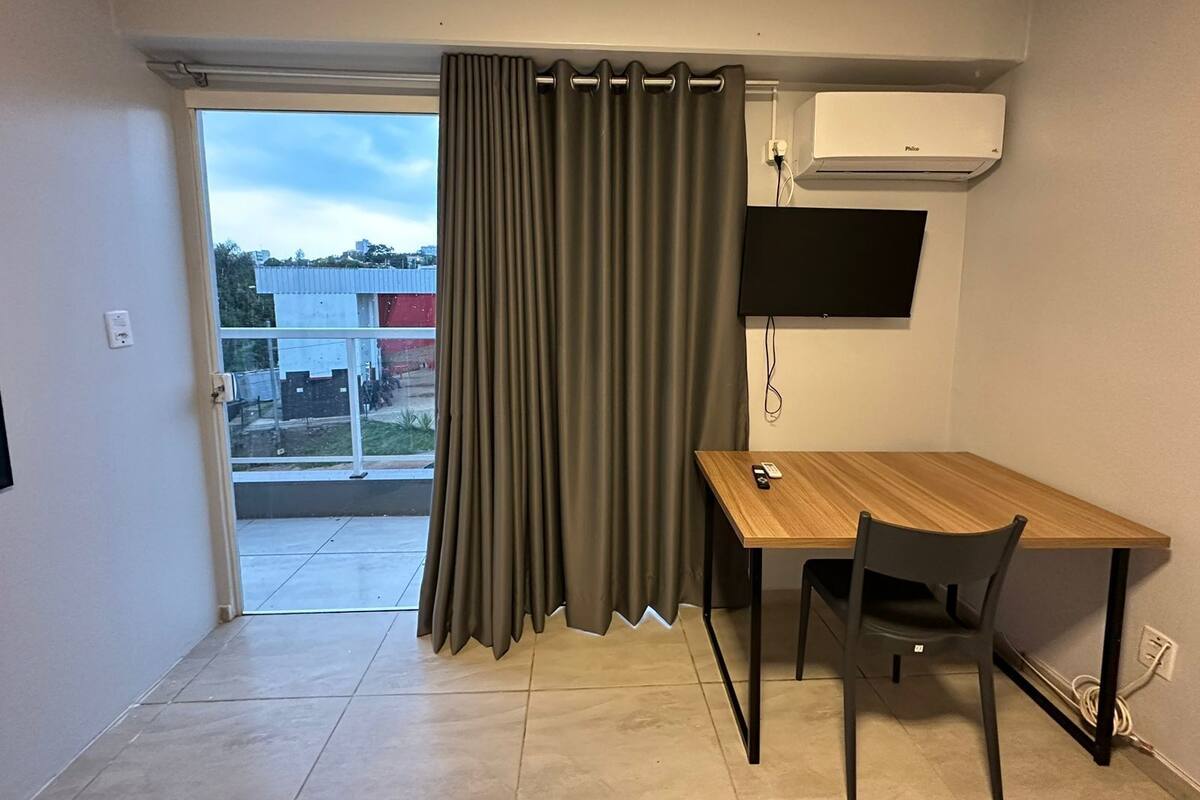 Um quarto pequeno com uma mesa de madeira e uma cadeira, uma TV presa na parede e uma janela com cortinas.