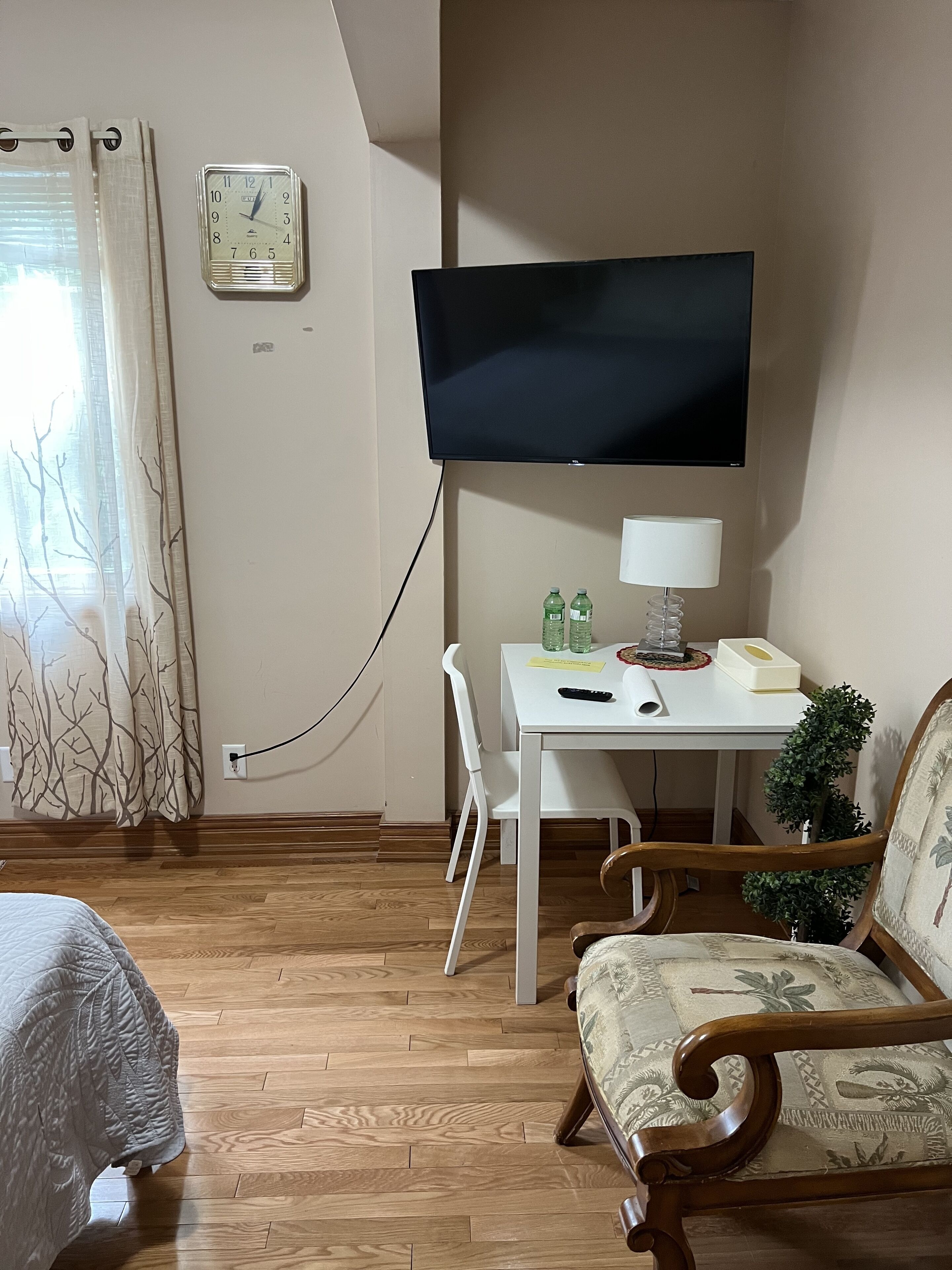 Quarto com uma cama, escrivaninha, cadeira, televisão, abajur e relógio na parede.
