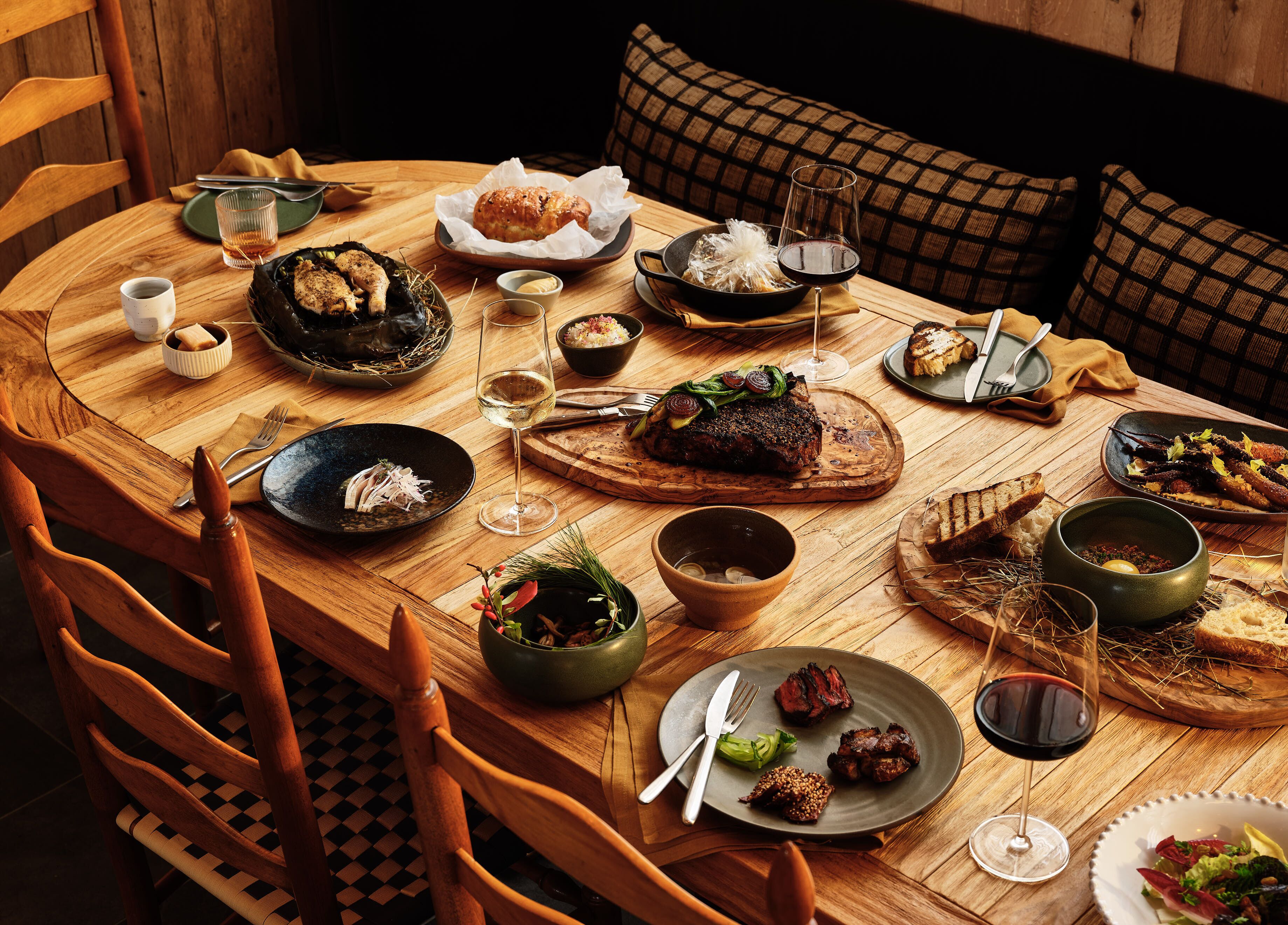 Uma mesa de jantar de madeira com diversos pratos, incluindo um centro de carne assada, rodeada por tigelas, pratos e copos de vinho.