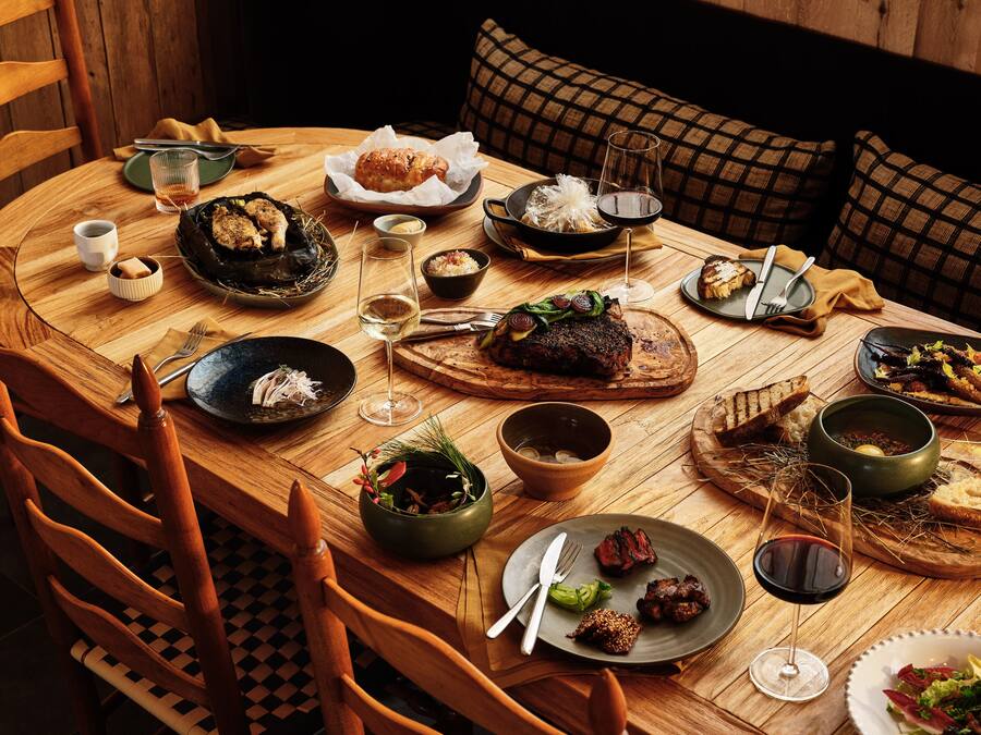 Uma mesa de jantar de madeira com diversos pratos, incluindo um centro de carne assada, rodeada por tigelas, pratos e copos de vinho.
