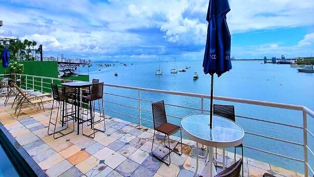 Um terraço com mesas ao ar livre, vista para o mar e um cais distante.