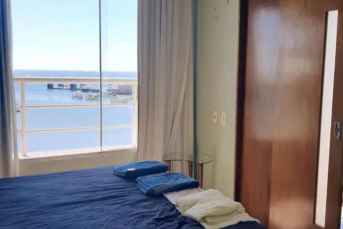 Quarto com cama, janela com vista para um corpo d'água e edifícios, porta e pequena mesa com abajur.