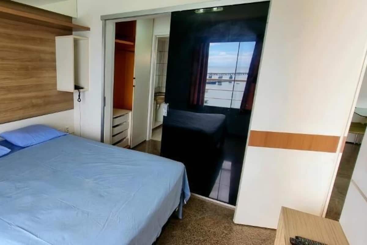 Quarto de hotel com uma cama grande, cabeceira de madeira, televisão, escrivaninha e vista para o mar pela janela.