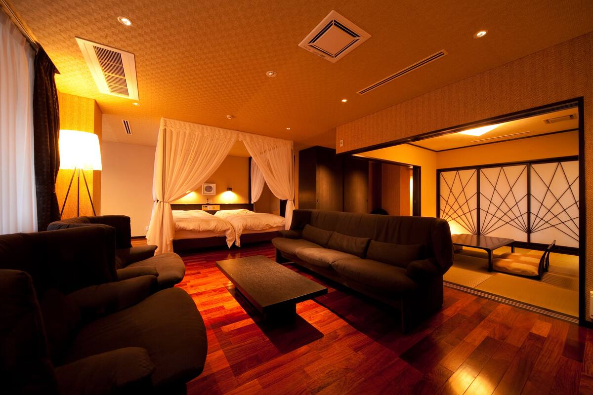 Quarto de hotel moderno com cama, sofá, mesa de centro e área de estar com uma mesinha baixa e almofadas.