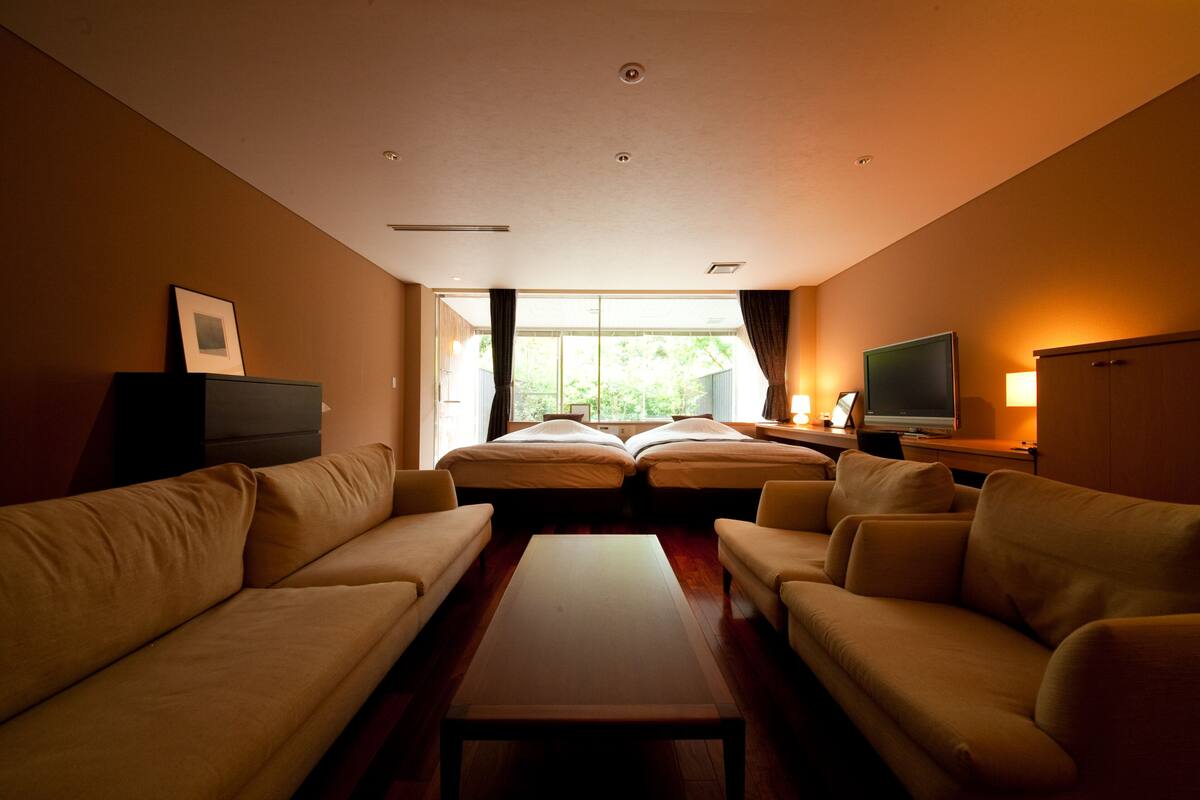 Quarto de hotel com duas camas de solteiro, um sofá, uma TV e uma escrivaninha com abajur.