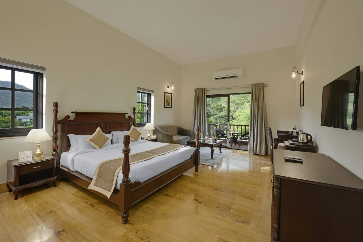 Um quarto com uma cama de dossel de madeira, uma mesa de cabeceira com luminária, uma escrivaninha com cadeira e uma janela grande com vista para o verde.