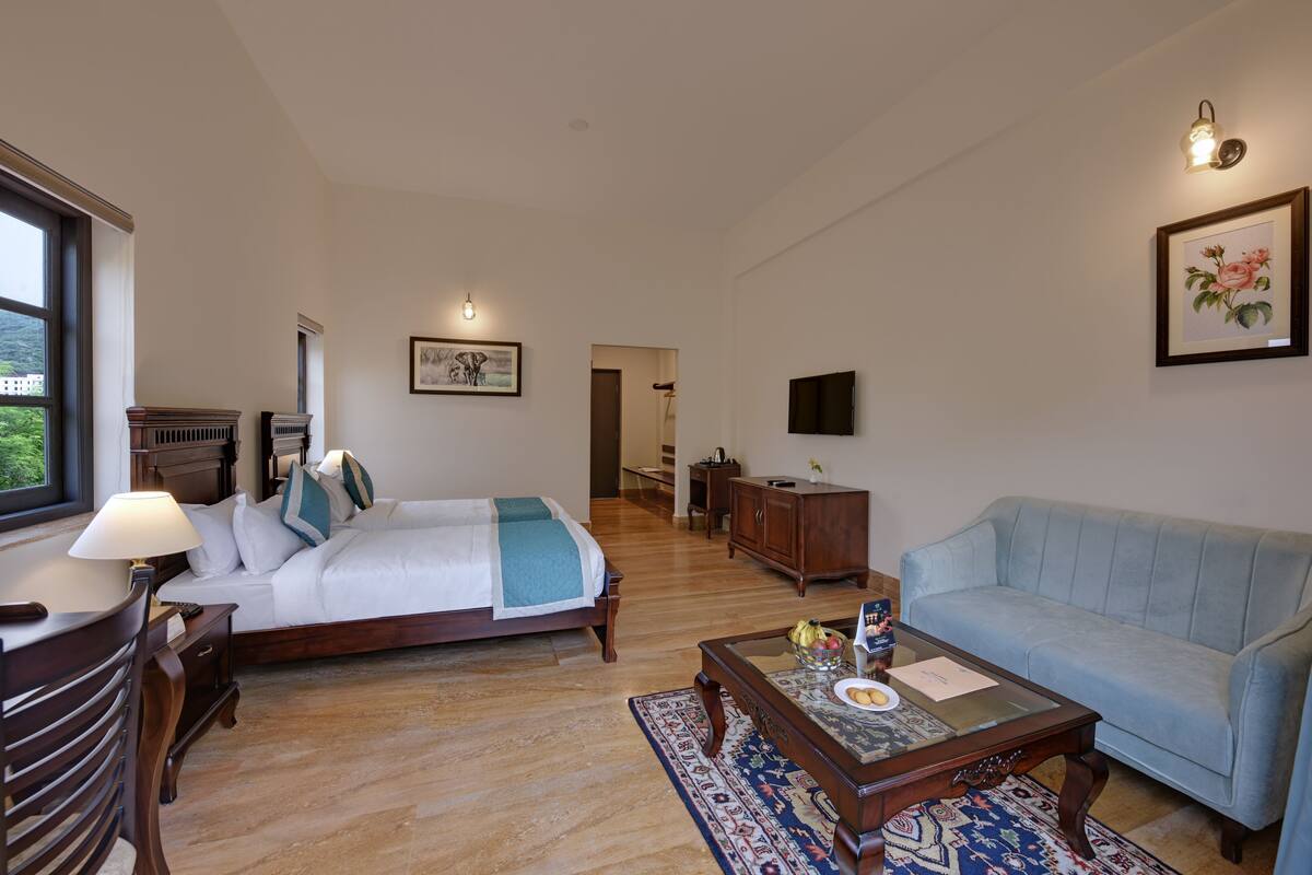 Quarto de hotel com cama, sofá, mesa de centro, escrivaninha com luminária, televisão e janela com vista.
