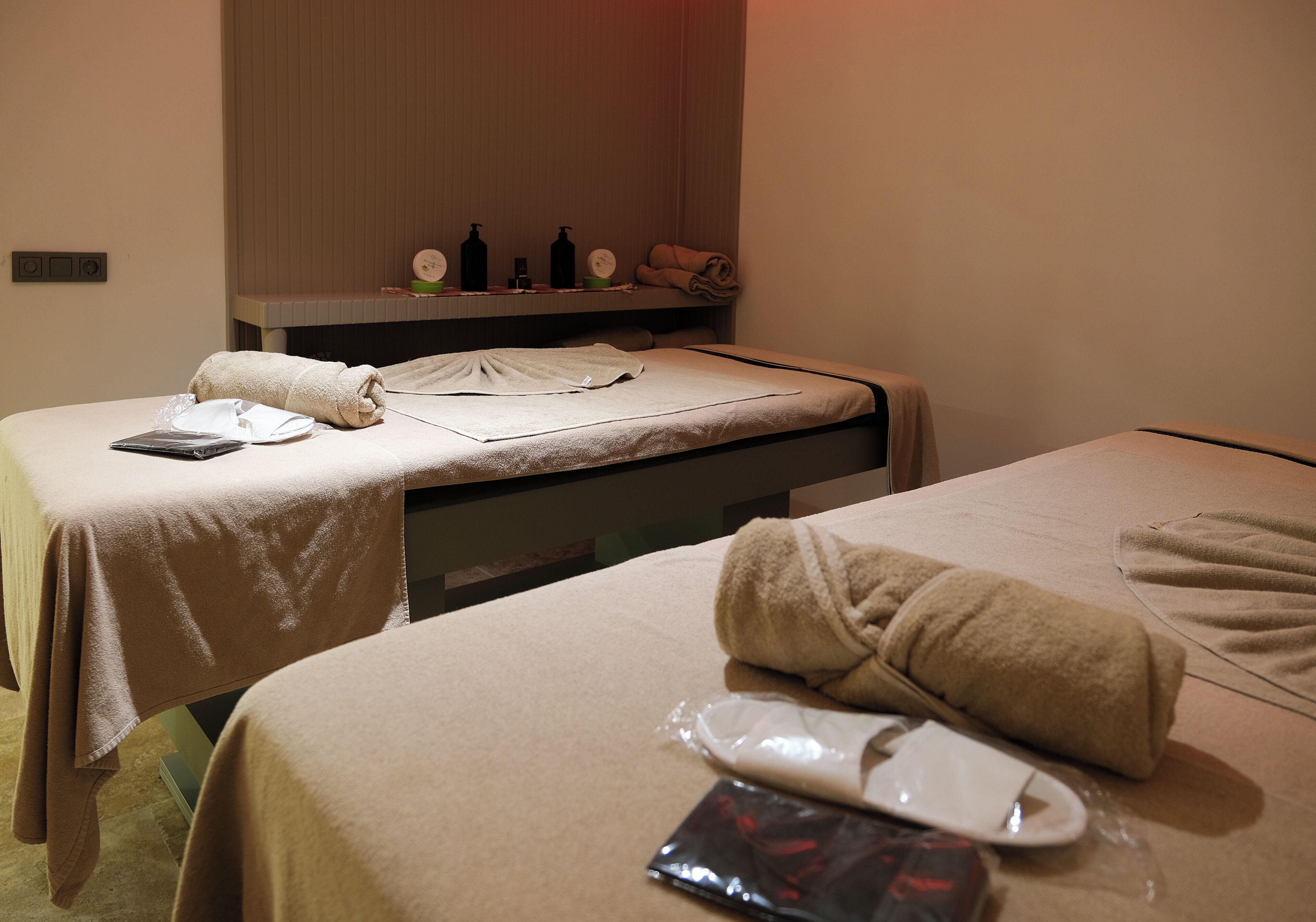 Quarto com duas camas, cada uma coberta por um cobertor bege, e uma pequena mesa com produtos de higiene pessoal.
