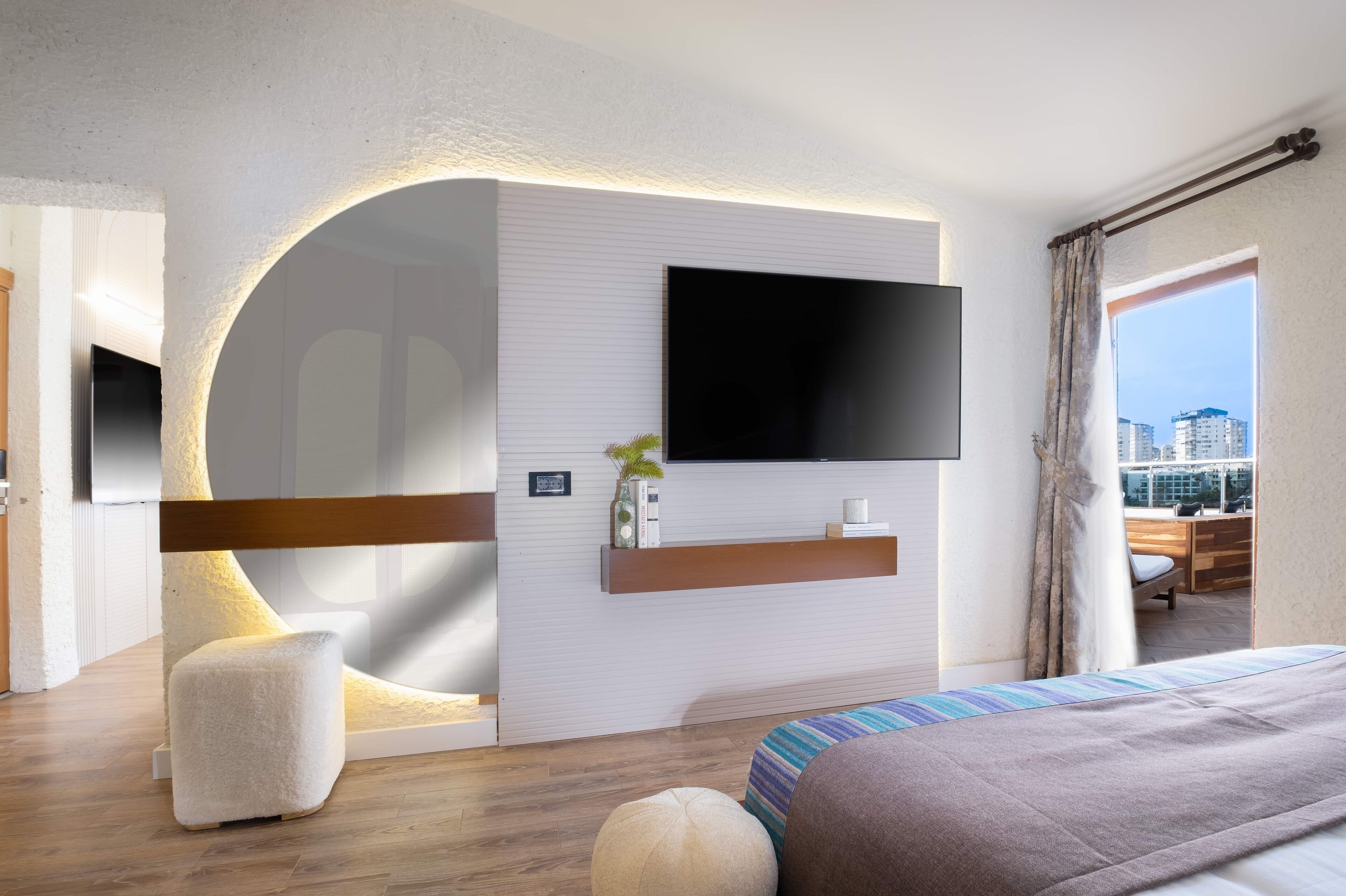 Quarto de hotel moderno com uma televisão de tela plana grande presa na parede, uma cama de madeira, um pequeno espaço de estar e vista para a cidade através de uma janela.