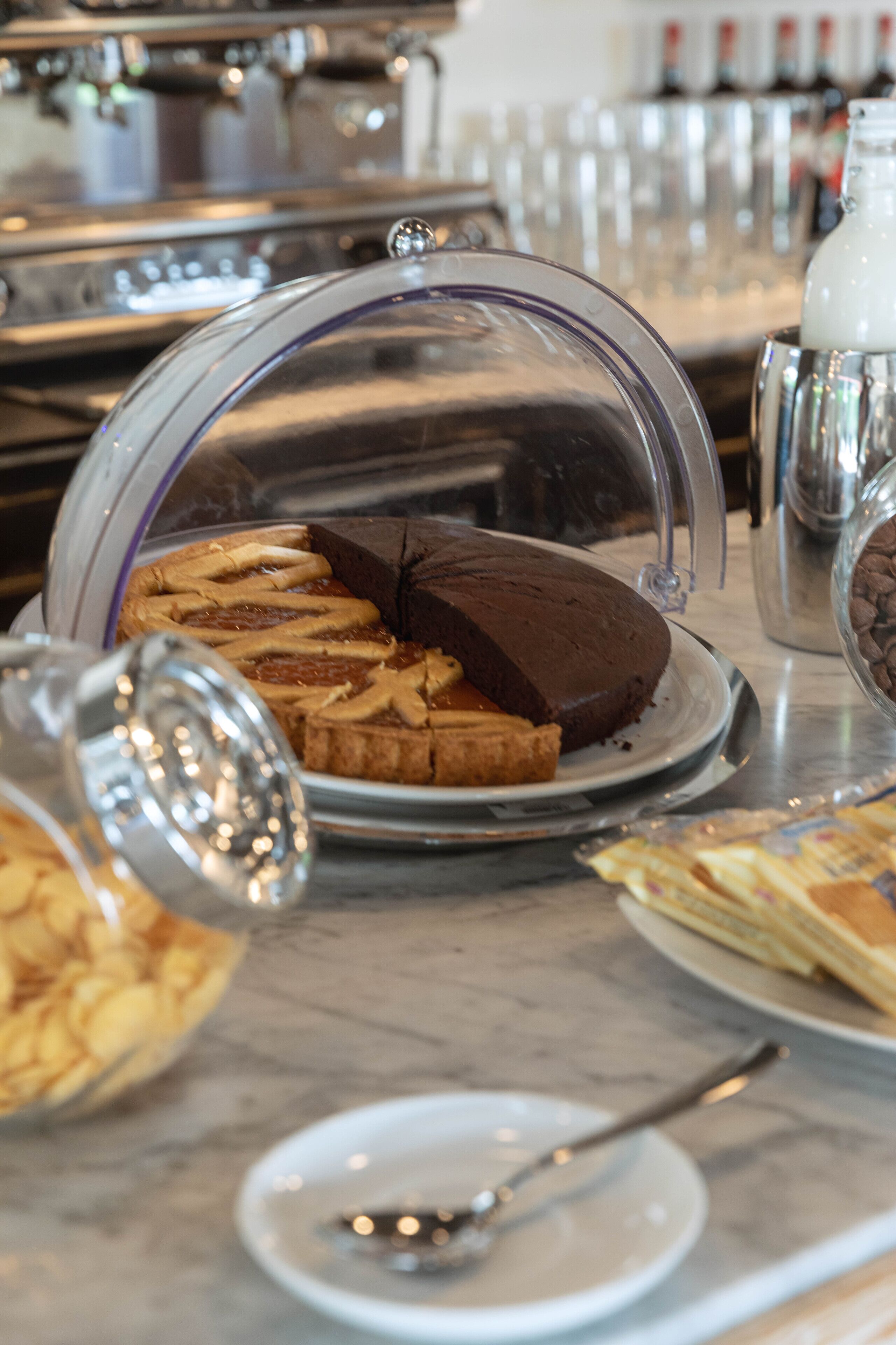 Exposição de sobremesas com um bolo de chocolate, uma cúpula de vidro e uma garrafa de molho sobre um balcão de mármore.