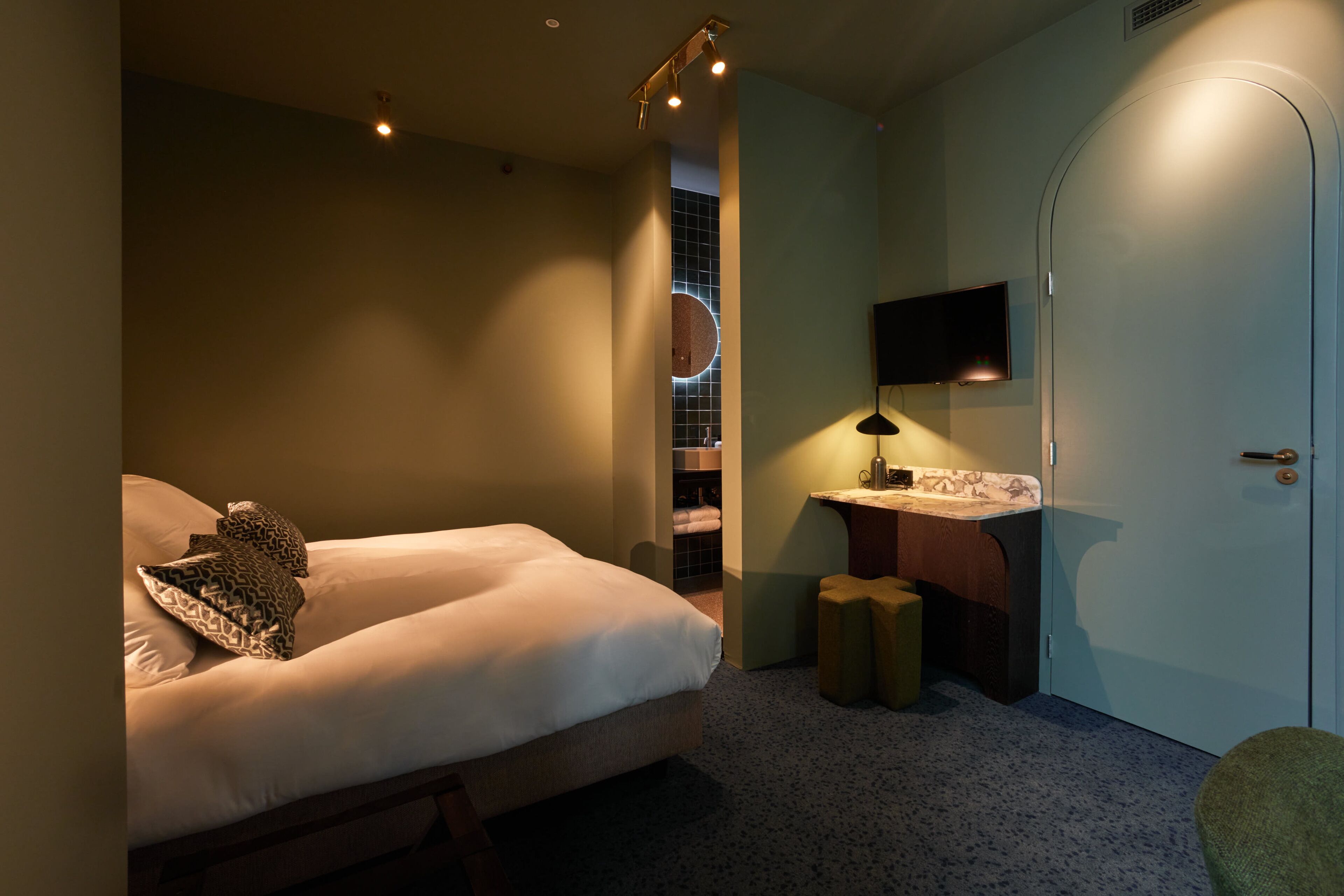 Quarto de hotel com cama, escrivaninha, televisão e porta.
