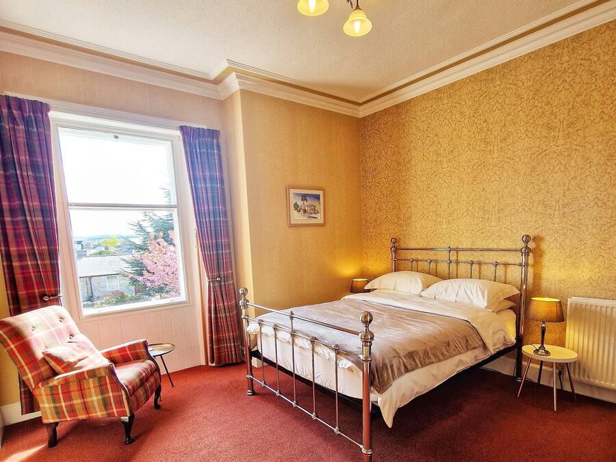 Quarto de hotel com cama, uma cadeira, uma mesinha, janela com cortinas e um candelabro.