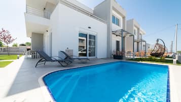 Uma área de piscina moderna com piscina azul cristalina, duas espreguiçadeiras e um edifício branco com grandes janelas e varanda.