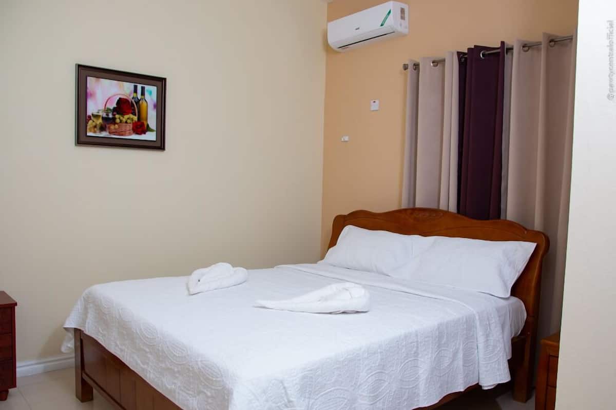 Cama bem arrumada com lençóis brancos e duas toalhas, ar condicionado fixado na parede e um quadro com uma cesta de frutas.