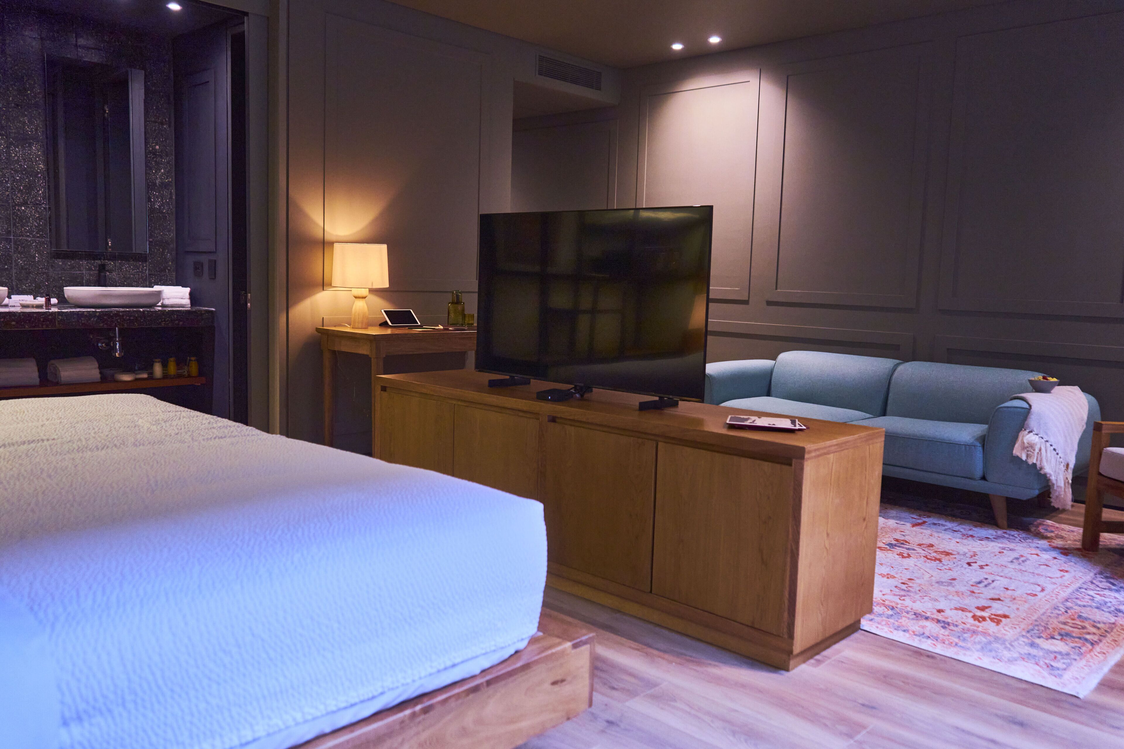 Quarto de hotel com cama, televisão sobre uma cômoda de madeira, sofá e banheiro com parede revestida de azulejos escuros e espelho.