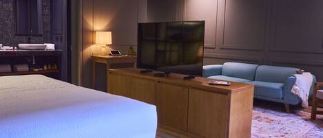 Quarto de hotel com cama, televisão sobre uma cômoda de madeira, sofá e banheiro com parede revestida de azulejos escuros e espelho.