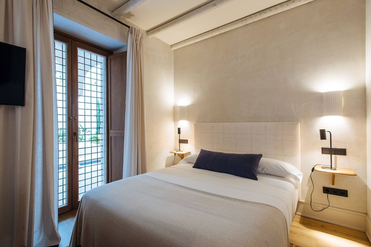 Quarto de hotel com cama, cortinas, televisão, mesa de cabeceira com abajur e janela com persianas.