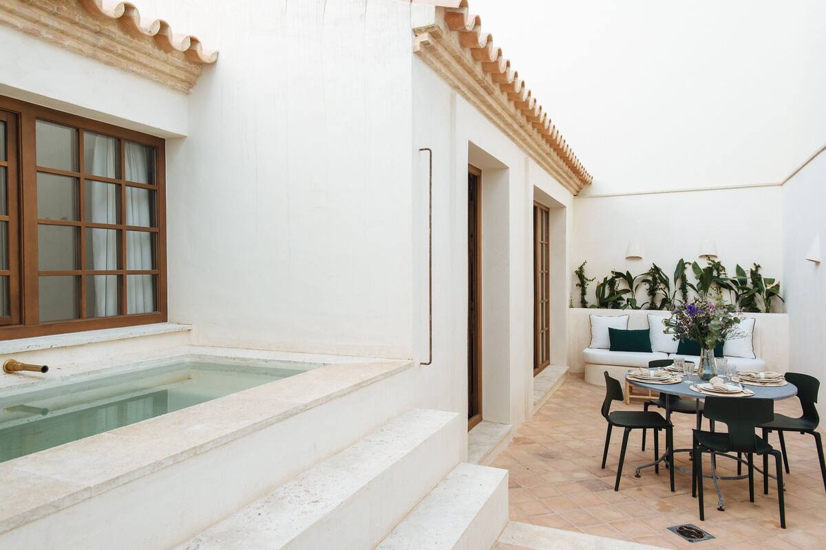 Área de piscina com degraus que levam a uma parede branca, uma mesa de jantar com cadeiras e um sofá com almofadas.
