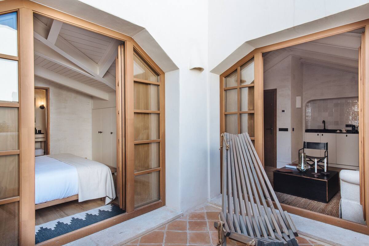 Quarto com uma cama, uma cadeira de madeira e um banheiro visível por uma porta aberta.