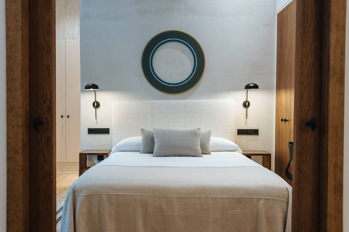 Quarto bem arrumado com uma cama grande, duas mesas de cabeceira, um adorno circular na parede e portas de madeira.