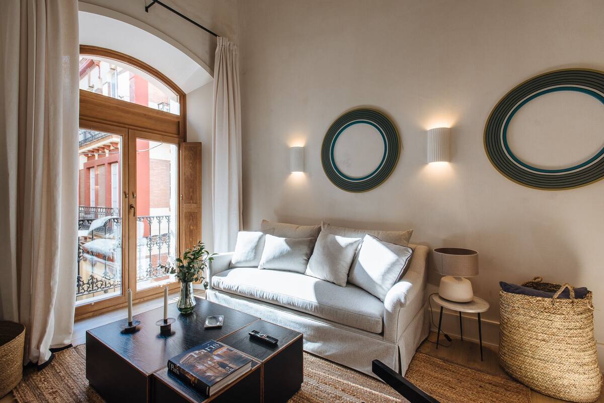 Uma sala de estar aconchegante com um sofá, uma mesa de centro com livros e um controle remoto, uma luminária e uma cesta grande de palha.