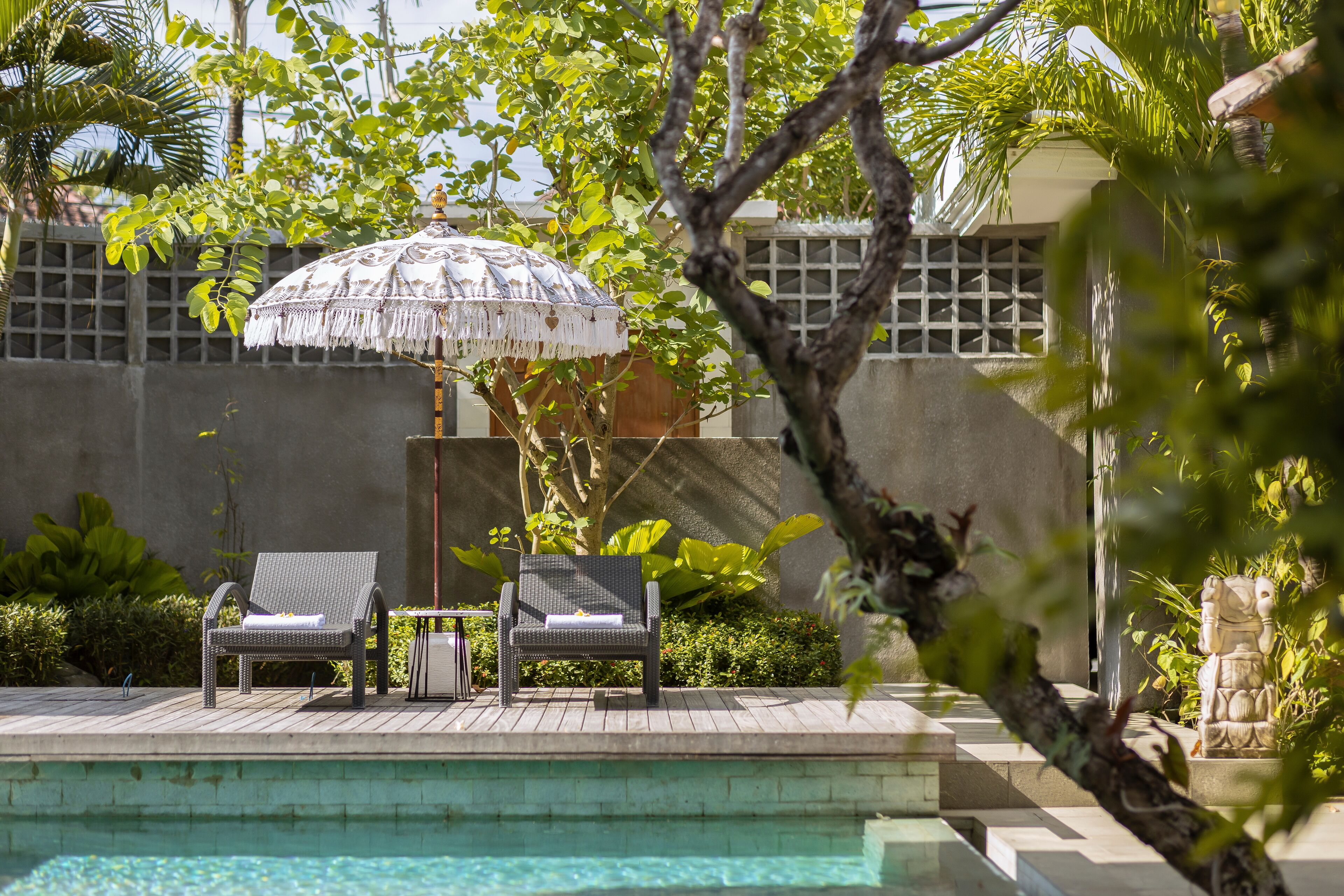 Uma área de piscina com duas espreguiçadeiras, um guarda-sol decorativo e uma estátua de pedra.