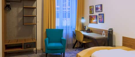Quarto de hotel com cama, secretária com cadeira, armário, janela com cortinas e radiador.