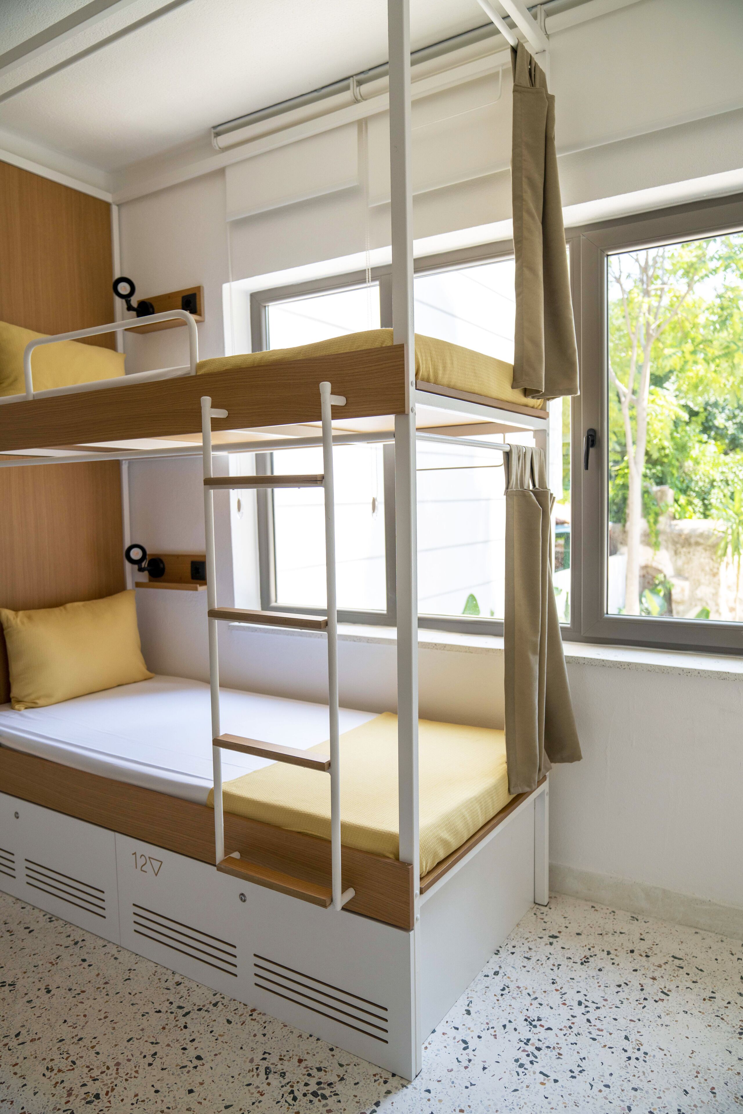 Uma cama beliche com escada de madeira, uma janela com cortina e uma pequena prateleira com luz de aro.
