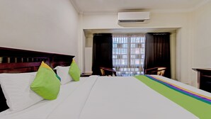 Quarto de hotel com uma cama grande, cabeceira de madeira, mesa de cabeceira, uma cadeira e uma janela com cortinas.