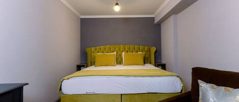 Quarto com cama estofada verde, duas mesas de cabeceira, piso de madeira e uma luminária suspensa.