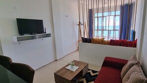 Uma sala de estar com uma televisão em uma estante, um sofá vermelho, uma mesa de centro de madeira com uma flor e uma janela com cortinas.