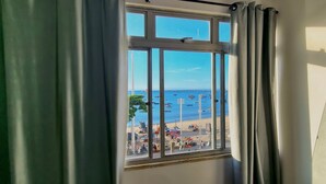 Janela com vista para uma praia, barcos e céu claro.