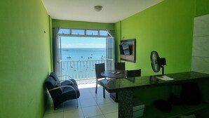 Quarto com paredes verdes, uma varanda com vista para o mar, uma mesa com cadeiras e um ventilador.