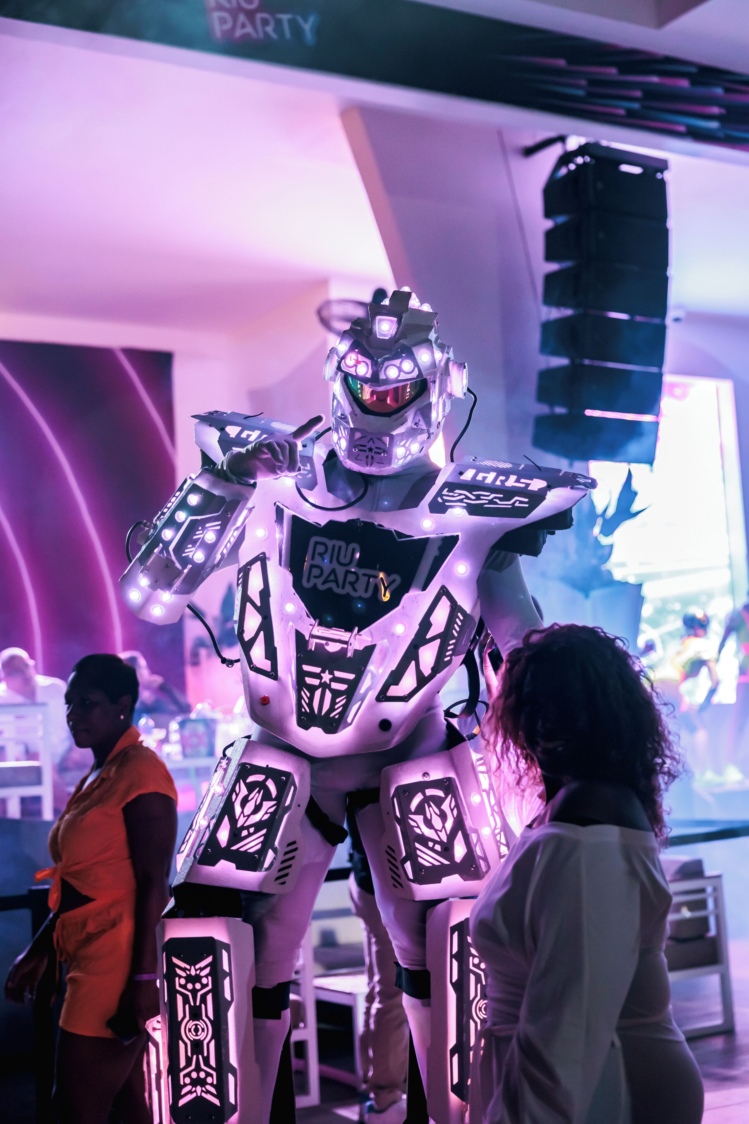 Uma pessoa vestida com um robô branco e preto com detalhes iluminados, em um quarto com tom roxo e um letreiro 'RIPARTY' ao fundo.