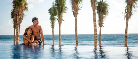 Um casal em uma piscina infinita privativa, com palmeiras e o oceano ao fundo.