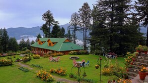 Uma hospedaria com telhado verde, rodeada por um jardim bem cuidado com diversas plantas e flores, e vista para montanhas ao fundo.