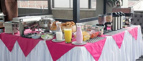 Um buffet de café da manhã com diversos alimentos e bebidas, disposto sobre uma mesa com toalha rosa e branca.
