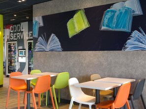 Um café moderno com cadeiras e mesas coloridas, um mural de livros na parede e uma máquina de vendas identificada como 'COOL SERVICE'.