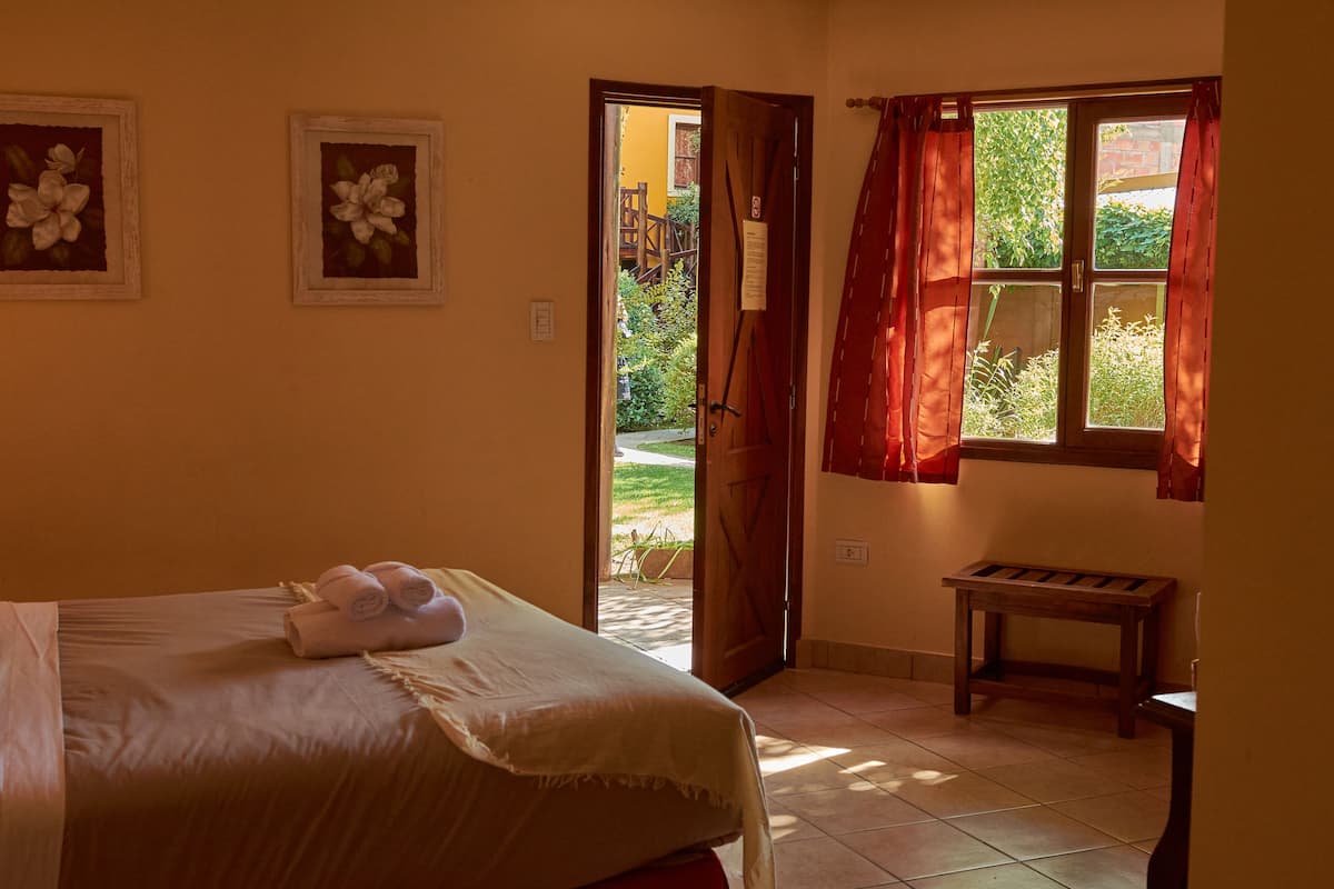 Quarto de hotel com cama, uma mesinha e janela com cortinas.