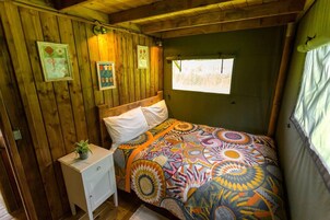 Um quarto com paredes de madeira, uma cama com colcha estampada e vibrante, uma pequena mesa de cabeceira com uma planta e quadros nas paredes.