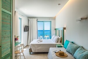 Quarto de hotel com uma cama, um sofá, uma mesinha com uma cadeira, uma televisão e vista para o mar pela janela.