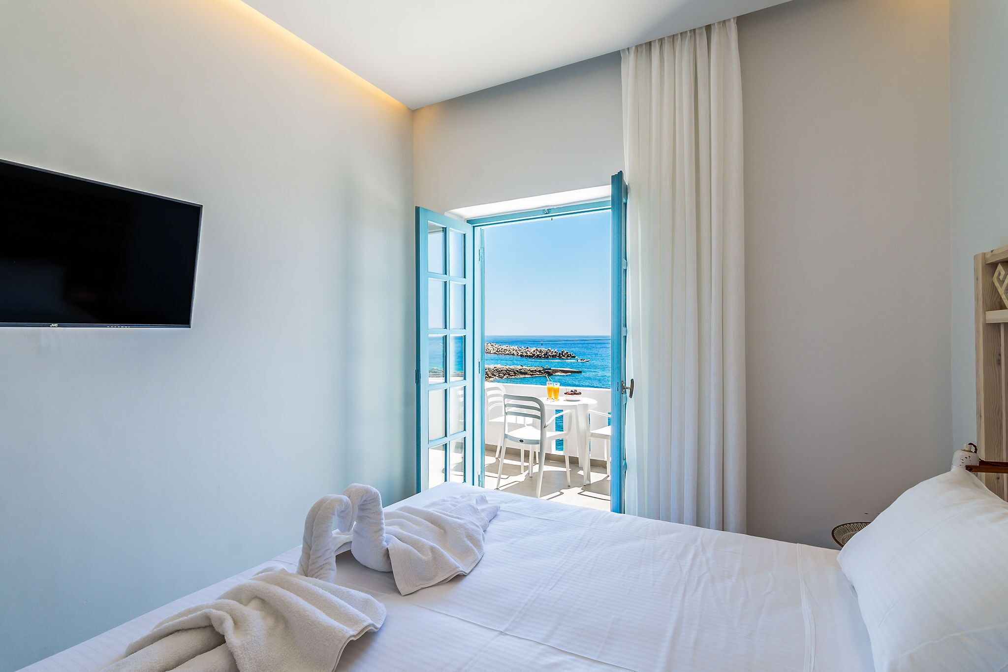 Quarto de hotel com uma cama, uma televisão na parede e uma porta aberta para uma varanda com vista para o mar.