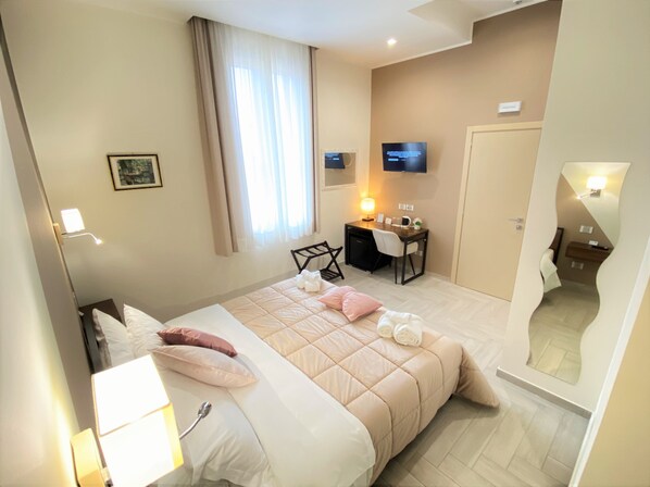 Quarto de hotel com cama, escrivaninha, cadeira, televisão e espelho no banheiro.