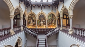 Uma escadaria imponente com corrimãos elaborados e uma série de arcos, que leva a uma galeria com pinturas grandes e detalhadas.